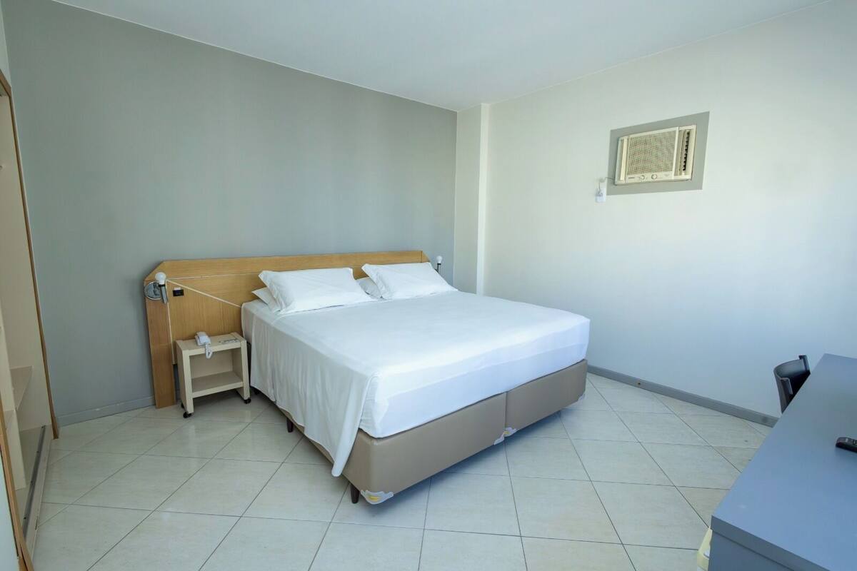 Quarto individual com uma cama de solteiro, cabeceira de madeira, mesa de cabeceira com telefone e escrivaninha com cadeira.