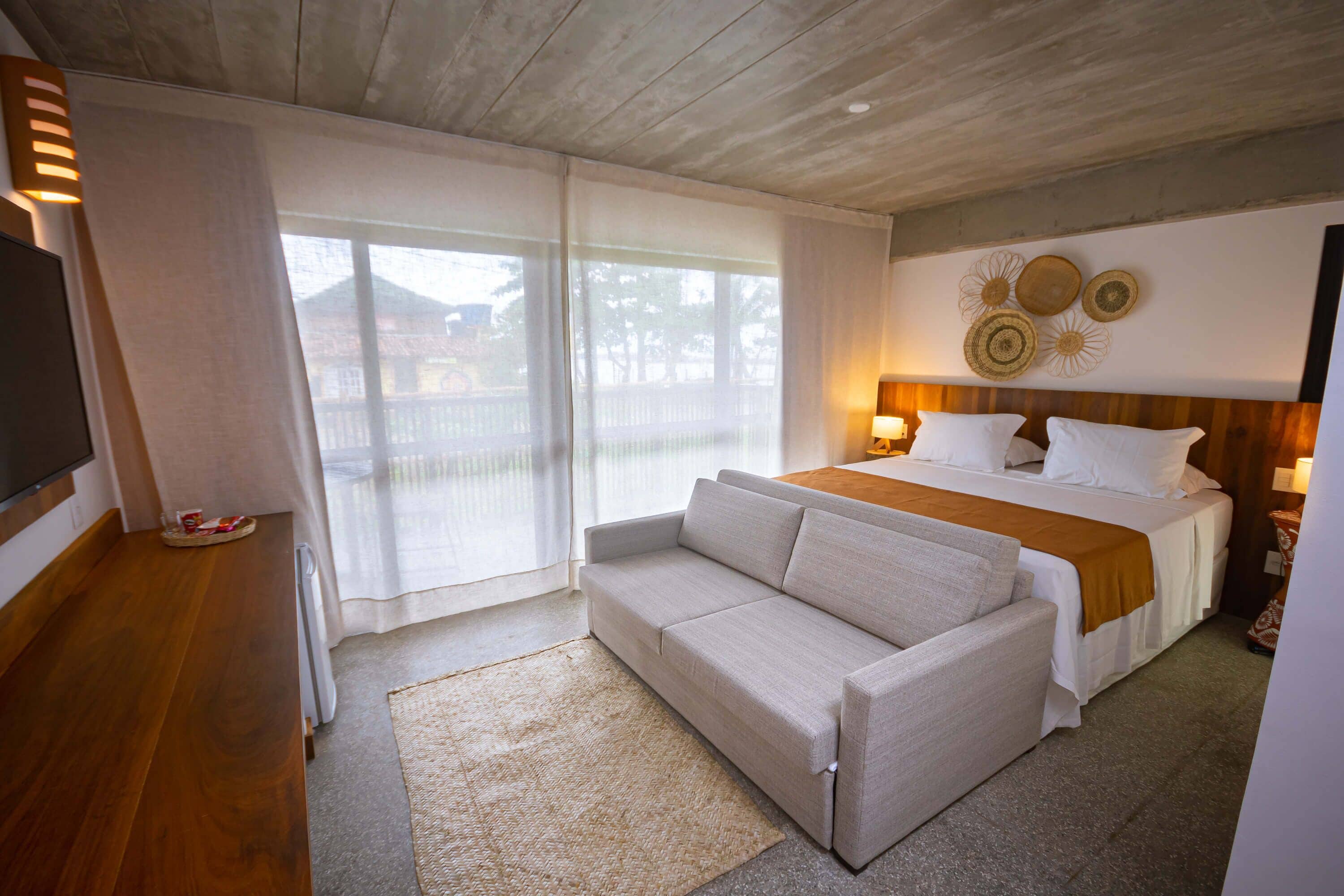 Quarto de hotel com cama, sofá, televisão e vista para o exterior.