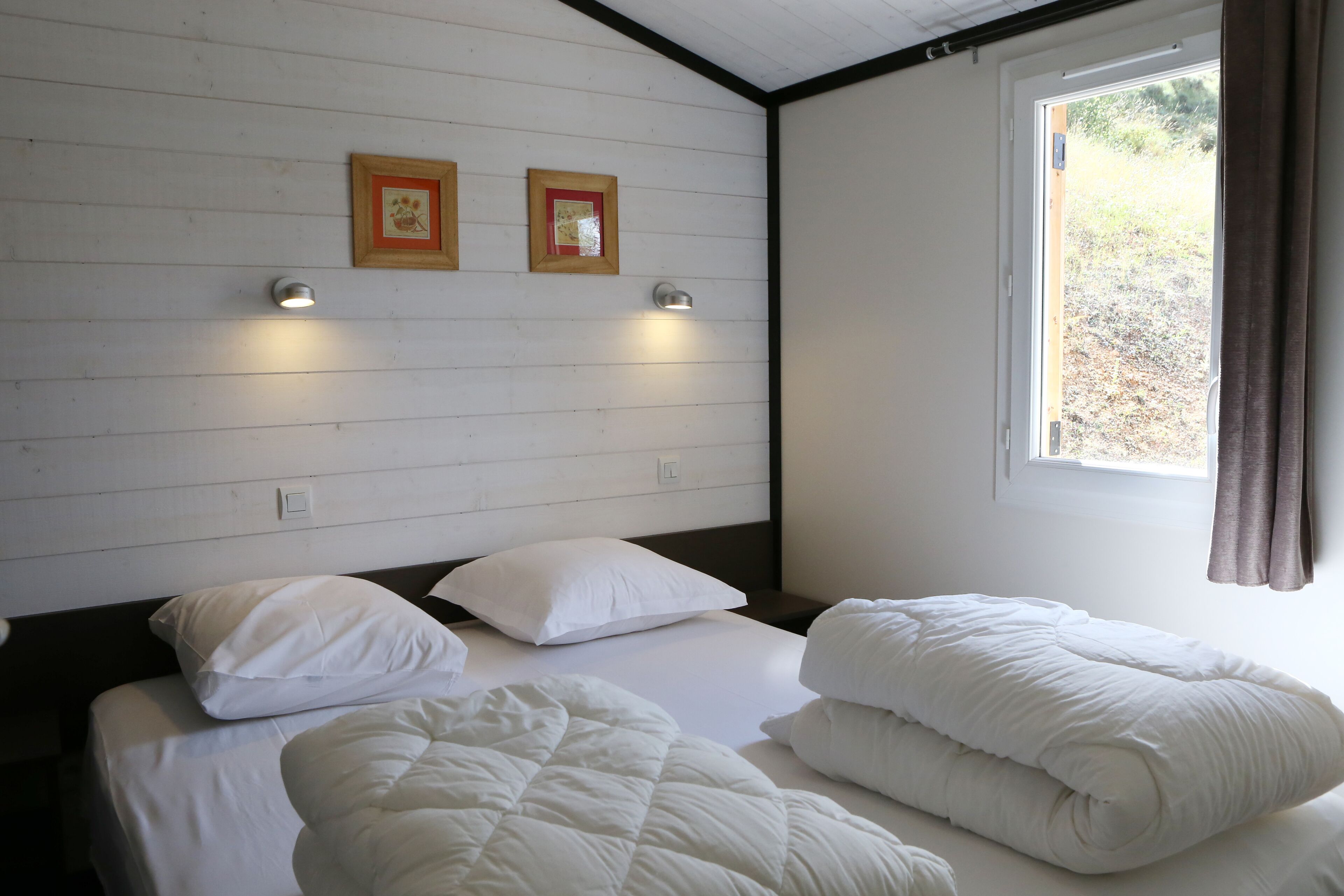 Quarto com duas camas, roupa de cama branca, janela com cortinas e luminárias na parede.