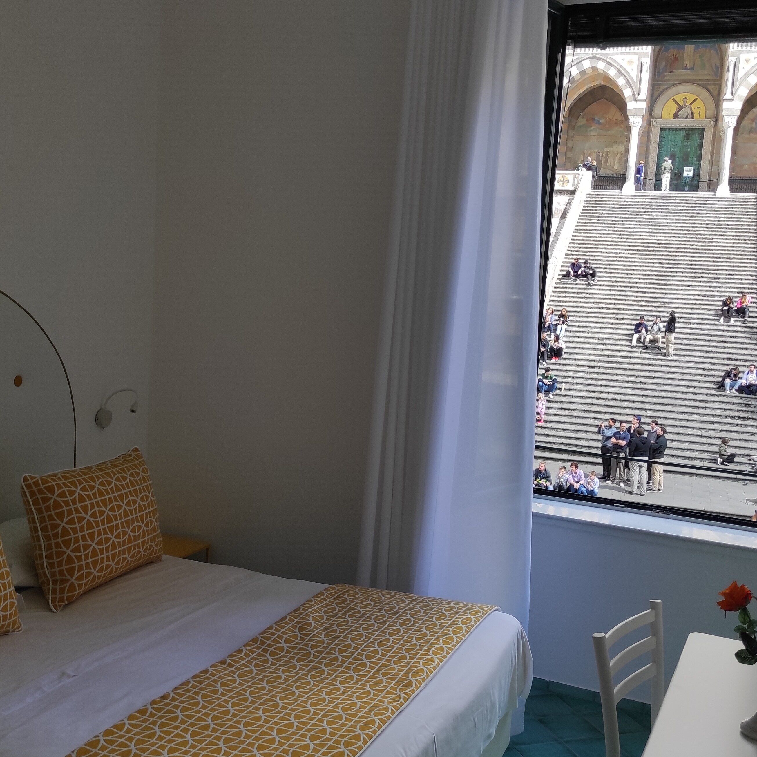 Quarto de hotel com cama, janela com cortinas, vista para uma praça pública com pessoas e uma mesa com um vaso de flores.