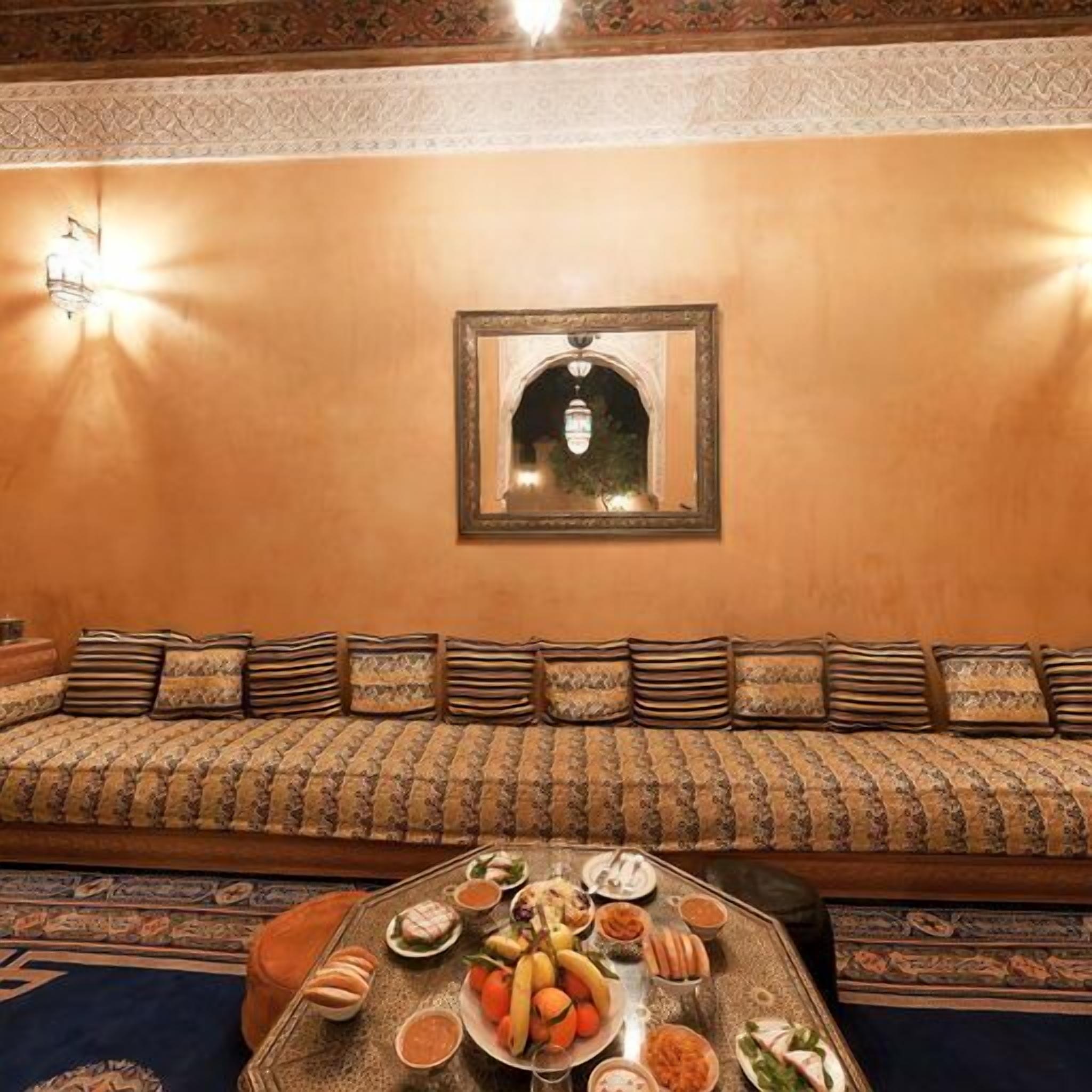 Um interior marroquino tradicional com um sofá estampado, uma mesa de jantar posta com diversos pratos e um espelho decorativo na parede.