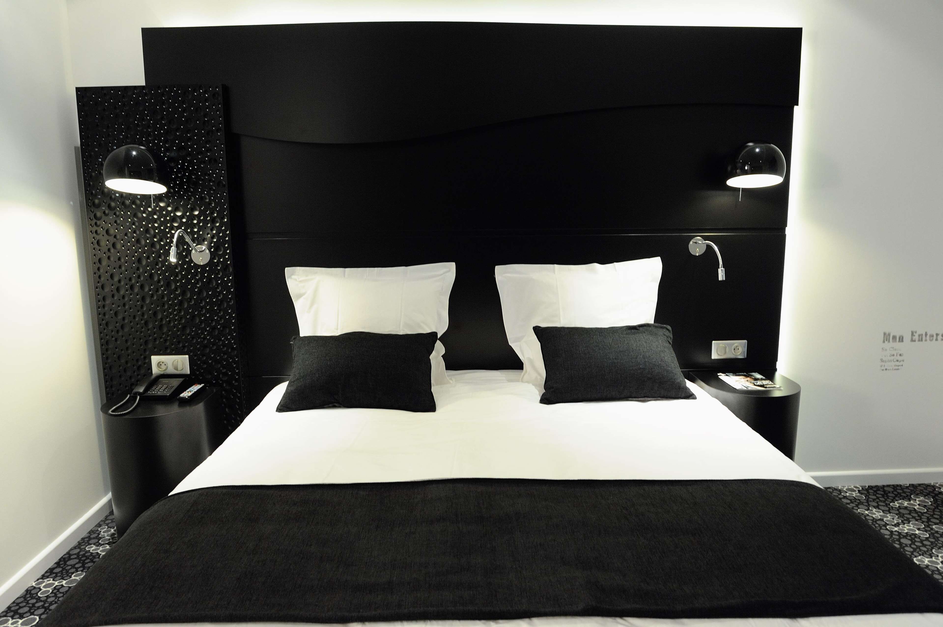Quarto de hotel moderno com uma cama grande, duas mesas de cabeceira, um painel de cabeceira preto e um candeeiro fixado na parede.