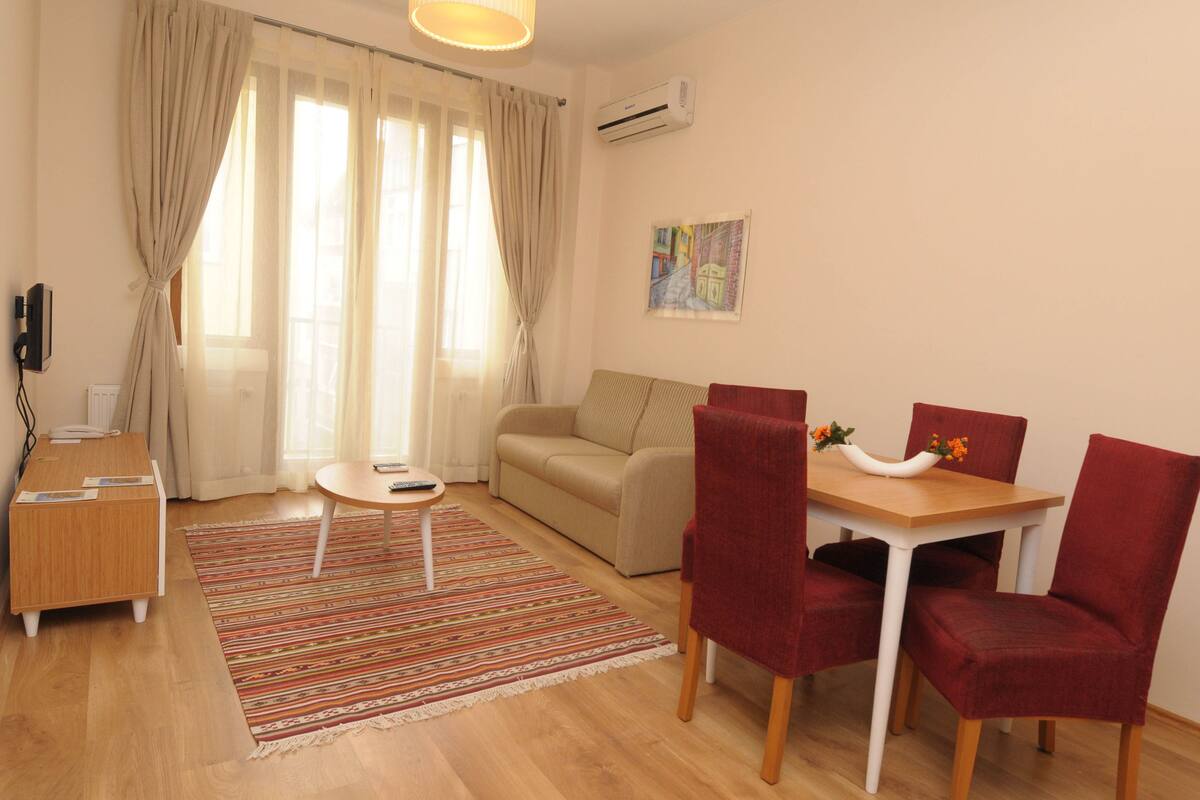 Uma sala de estar moderna com sofá, mesa de jantar com cadeiras, uma mesinha de centro, tapete estampado e um quadro na parede.