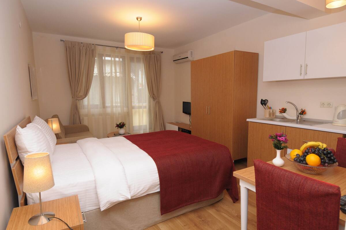 Quarto de hotel com cama, mesa de cabeceira, luminária, cortinas, uma pequena mesa com um vaso de flores, uma fruteira e uma cozinha compacta com armários e pia.