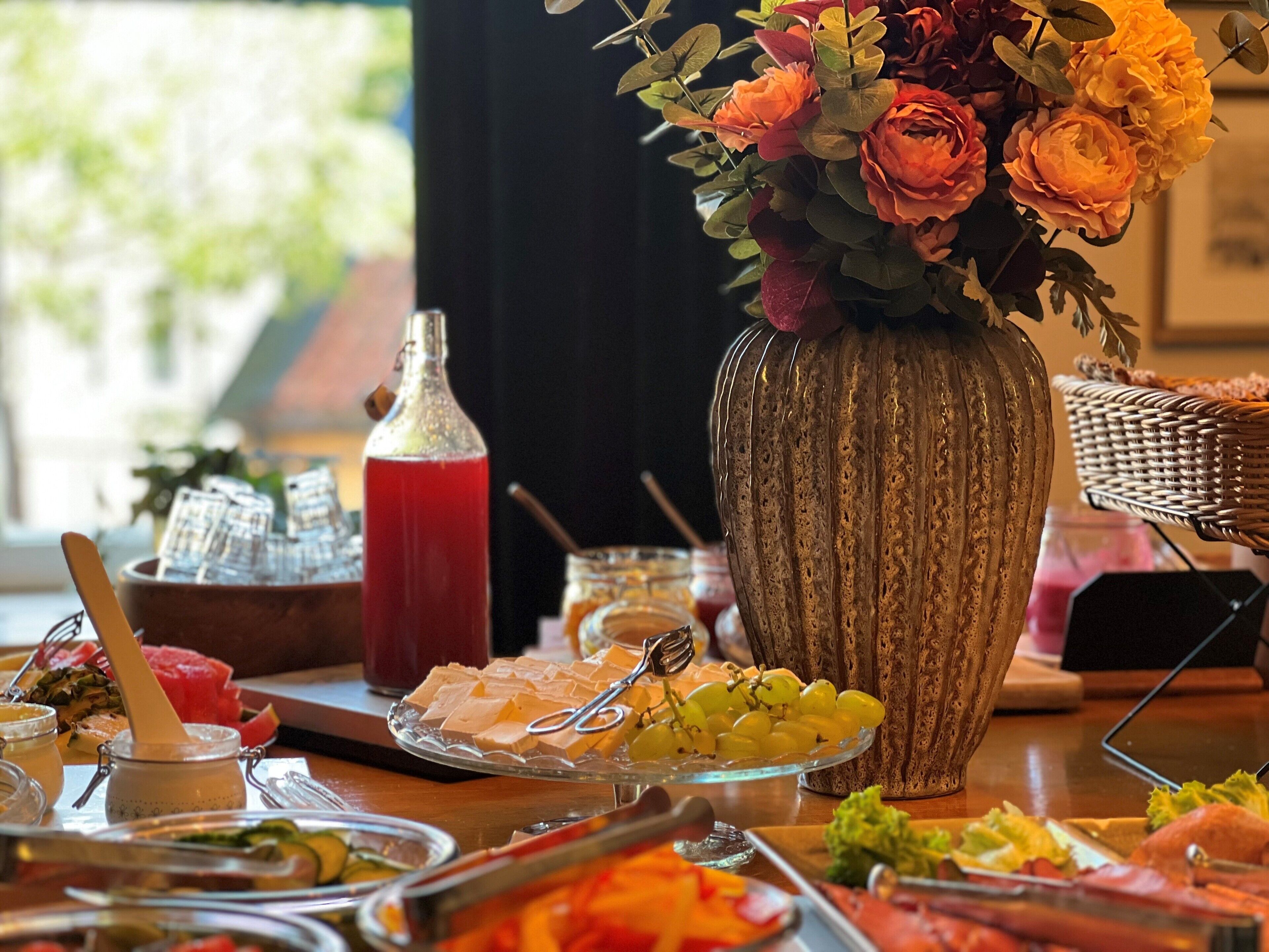 Uma mesa de buffet com diversos alimentos, um vaso decorativo com flores e uma garrafa de bebida vermelha.