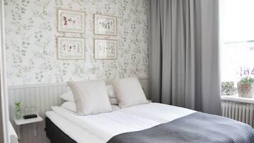 Cama bem arrumada com lençóis brancos e manta cinza, mesa de cabeceira com um vaso e copos, papel de parede floral e janela com cortinas.
