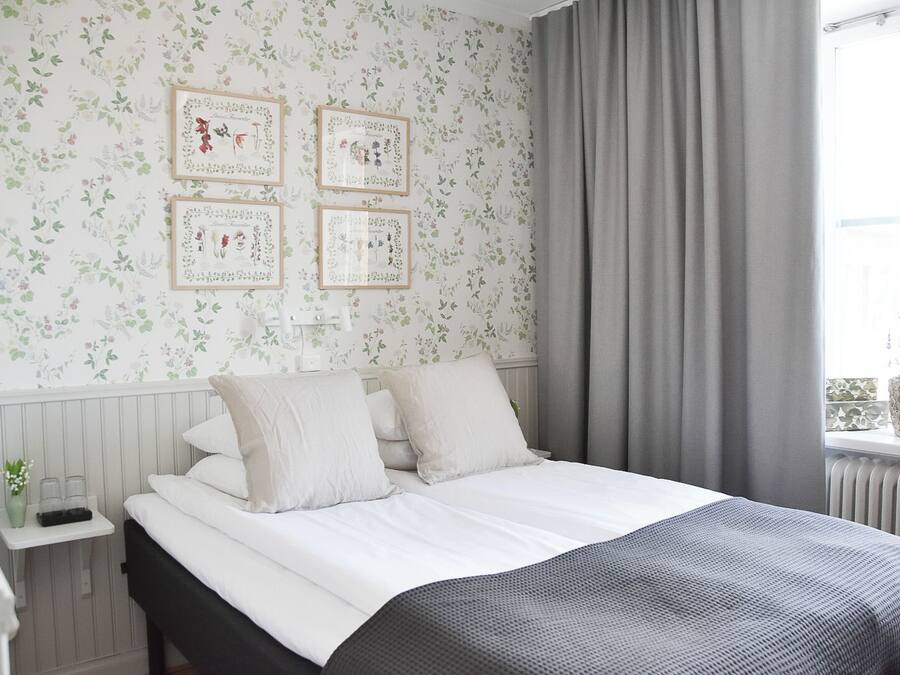 Cama bem arrumada com lençóis brancos e manta cinza, mesa de cabeceira com um vaso e copos, papel de parede floral e janela com cortinas.