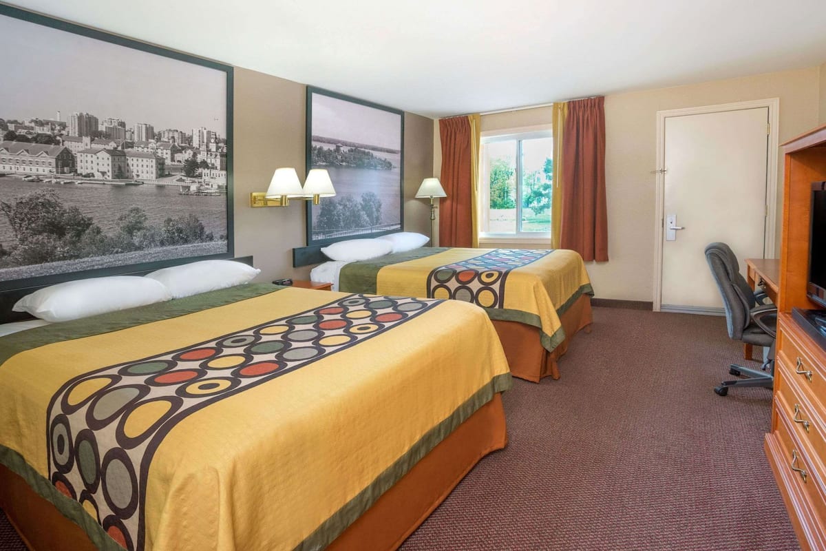 Quarto de hotel com duas camas, uma escrivaninha, uma cadeira, televisão e uma janela grande com cortinas.
