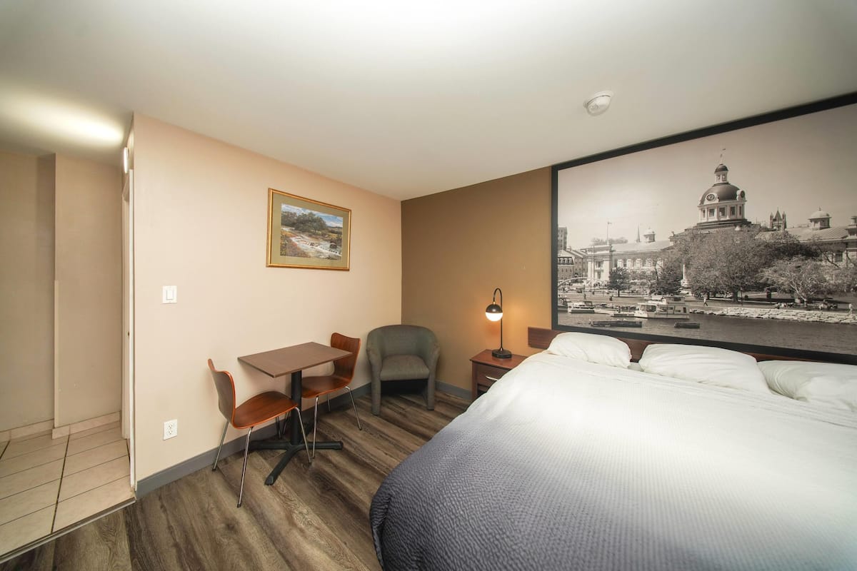 Quarto de hotel com uma cama, uma escrivaninha com cadeira, uma luminária, um quadro na parede e uma janela grande com vista para um edifício.