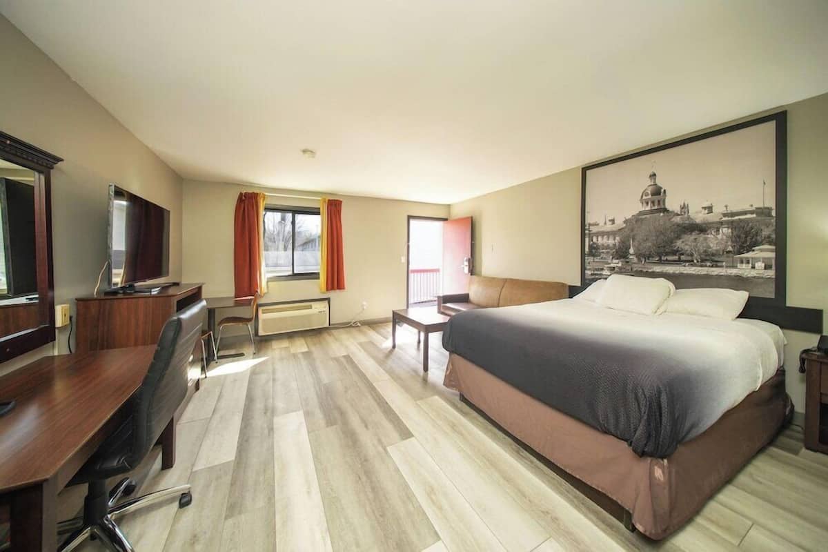 Quarto de hotel com cama, escrivaninha, cadeira e uma janela grande.
