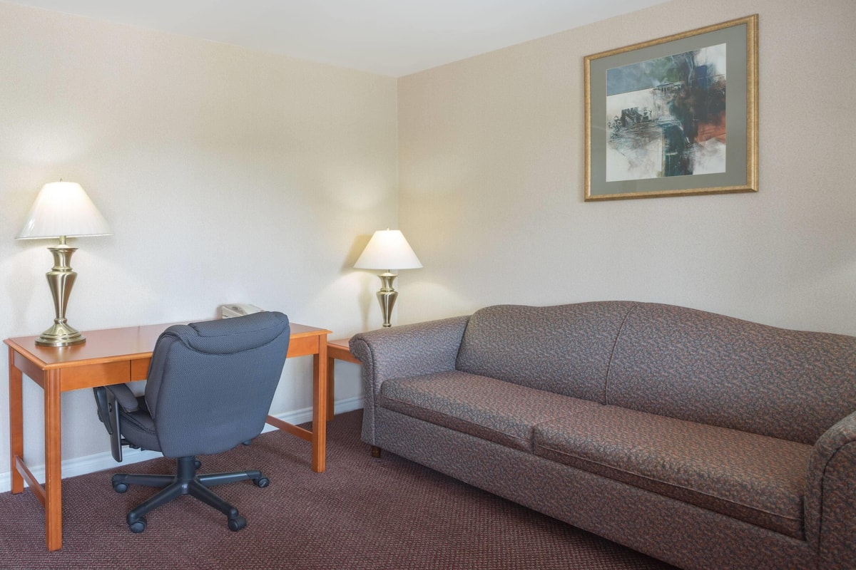 Quarto de hotel com escrivaninha de madeira, duas luminárias, uma cadeira de escritório cinza, um sofá castanho e um quadro abstrato emoldurado na parede.