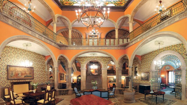 Um hall imponente com tetos ricamente decorados, um banquinho vermelho central e uma área de estar ampla com várias cadeiras e mesas.