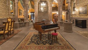 Um amplo hall de entrada com um piano de cauda, lustres ornamentados e uma escadaria para um andar superior.