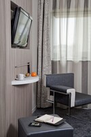 Quarto de hotel moderno com televisão de tela plana na parede, uma pequena prateleira branca com uma xícara, um kiwi e uma laranja, uma poltrona cinza estofada e um banquinho preto com um caderno e uma caneta.