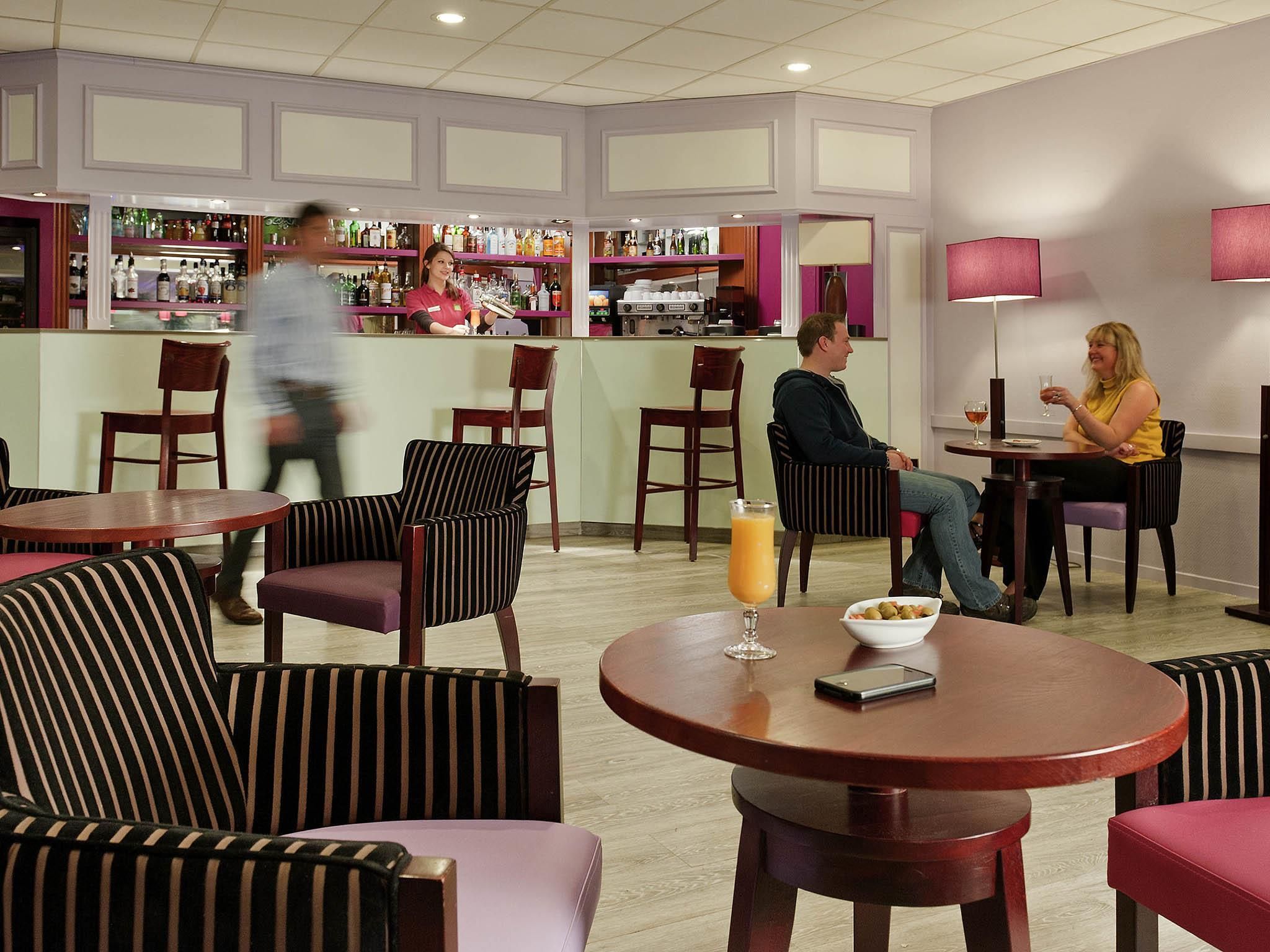 Uma área de bar com balcão e banquetas, um espaço de estar com mesas redondas e cadeiras listradas, e uma pessoa andando ao fundo.