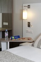 Quarto de hotel com cama, mesa de cabeceira de madeira, garrafa de água, cartão de boas-vindas e luminária fixada na parede.