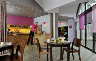 Um espaço de refeições moderno em hotel, com mobiliário de madeira, uma cozinha ao fundo e grandes janelas.