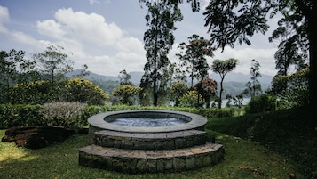 Uma banheira de pedra rodeada por vegetação exuberante e árvores, com montanhas ao fundo.