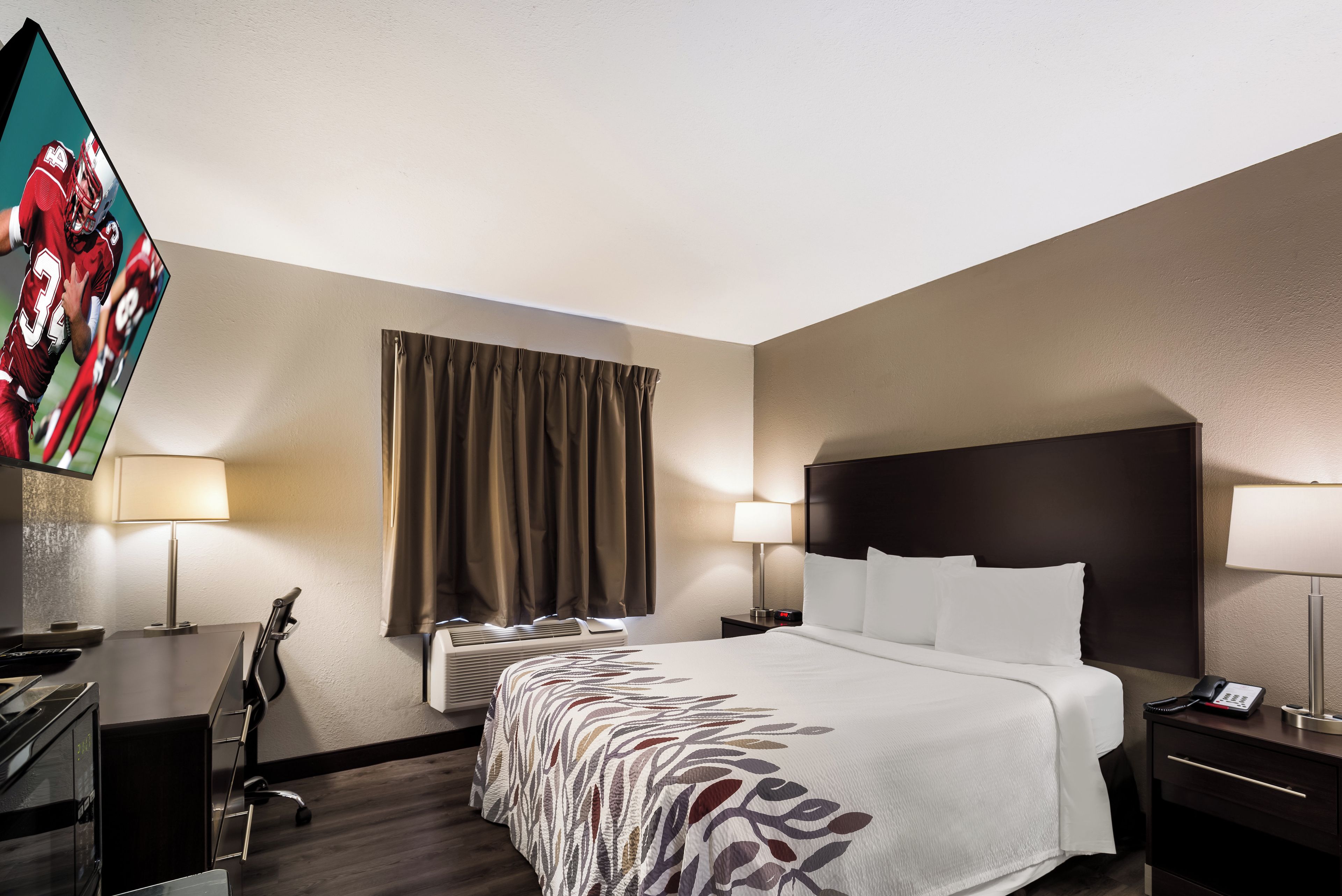 Quarto de hotel com cama, abajures, escrivaninha com computador, televisão e janela com cortinas.