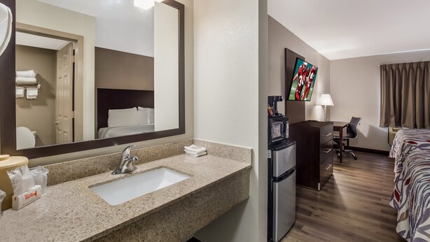 Quarto de hotel com pia de bancada de granito, espelho, frigorífico, secretária com cadeira e cama com roupa de cama estampada.
