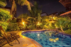 Piscina com bordas azulejadas, cercada por palmeiras e cadeiras de descanso.