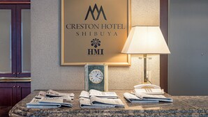 Bancada de recepção de hotel com uma placa emoldurada do Creston Hotel Shibuya, um relógio, uma luminária e jornais espalhados.