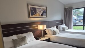 Quarto de hotel com duas camas, uma mesinha de cabeceira, abajur, um quadro e vista para o exterior.