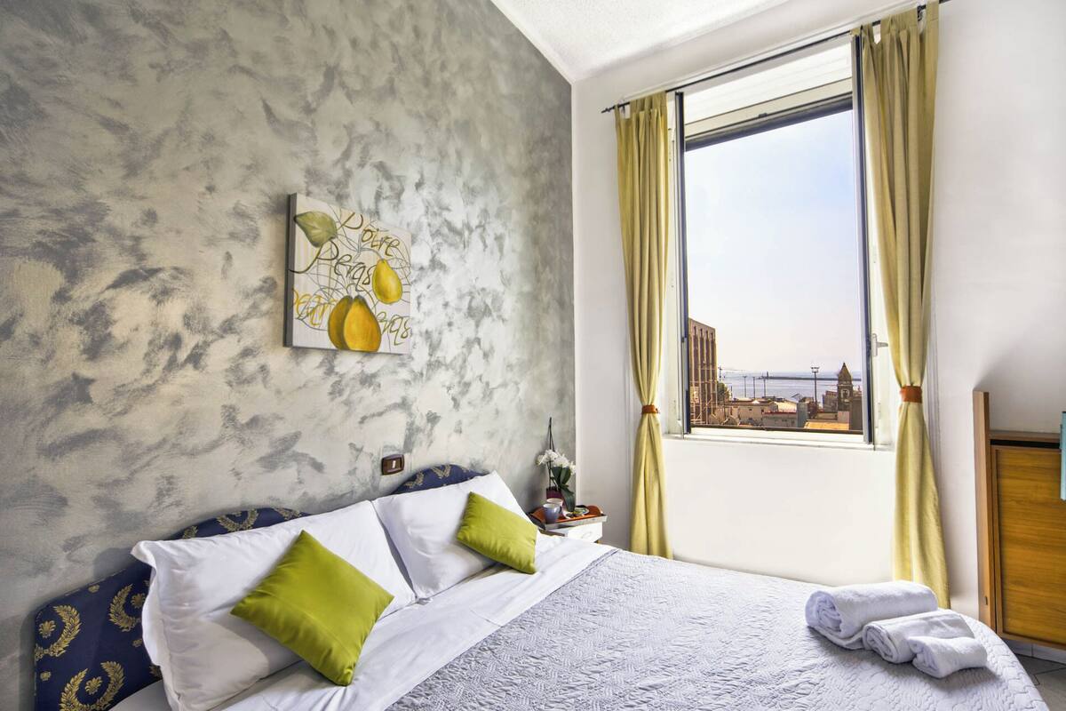 Quarto com cama, janela com cortinas, parede com pintura e vista da cidade.