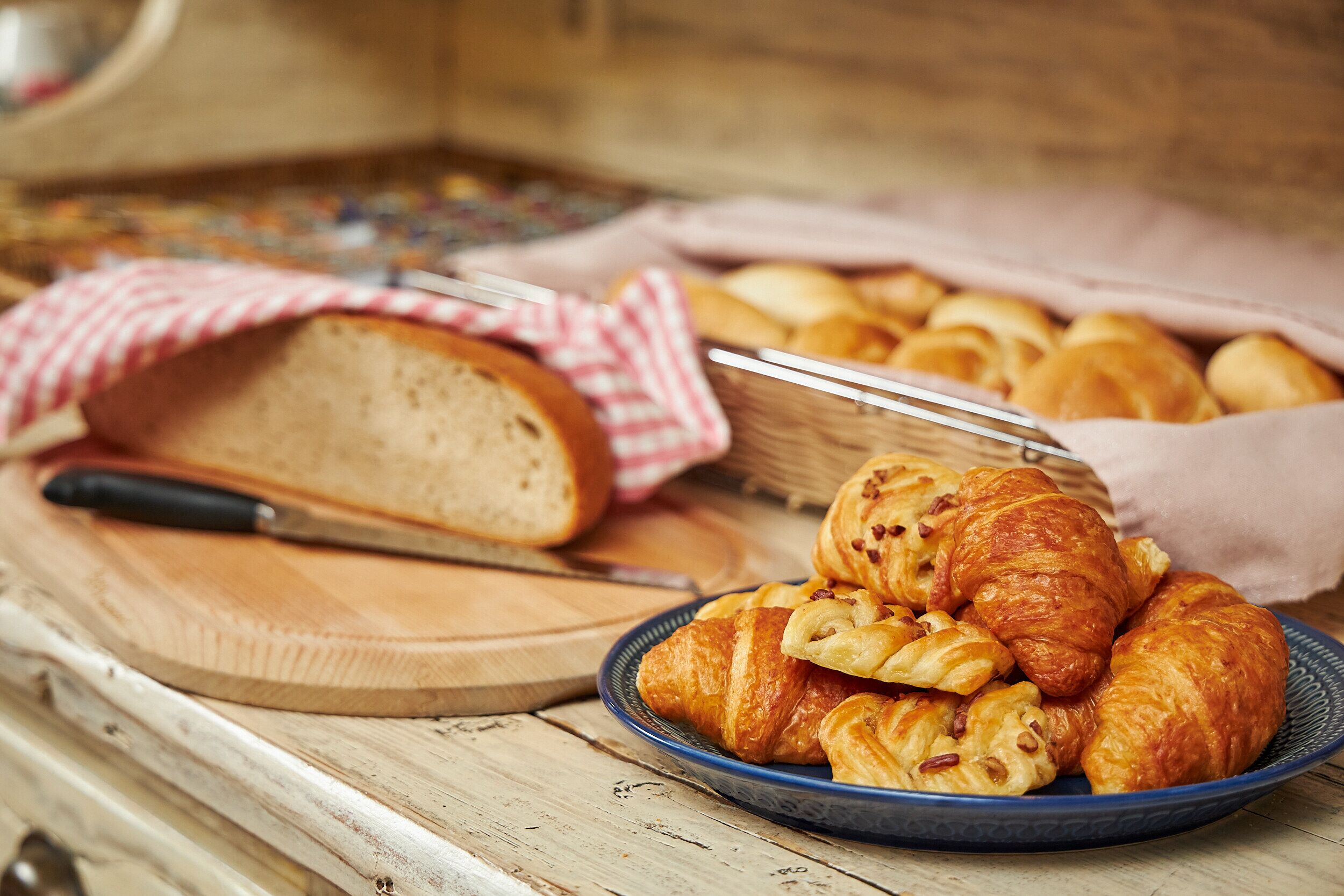 Tábua de madeira com um pão e uma faca, um prato azul com croissants, e uma cesta de pães.