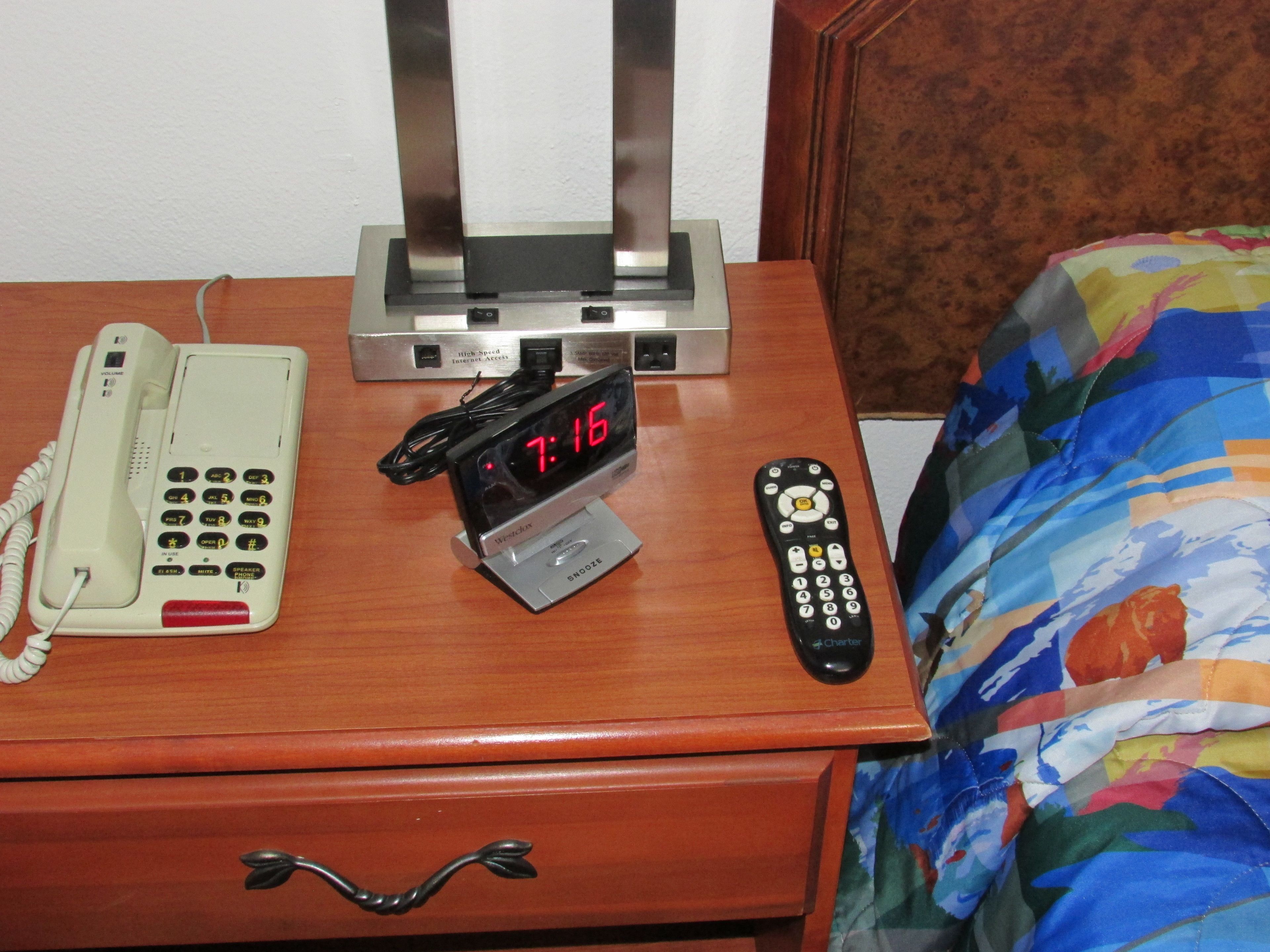 Uma mesa de cabeceira de madeira com um telefone branco, um relógio digital marcando 7:16 e um controle remoto.