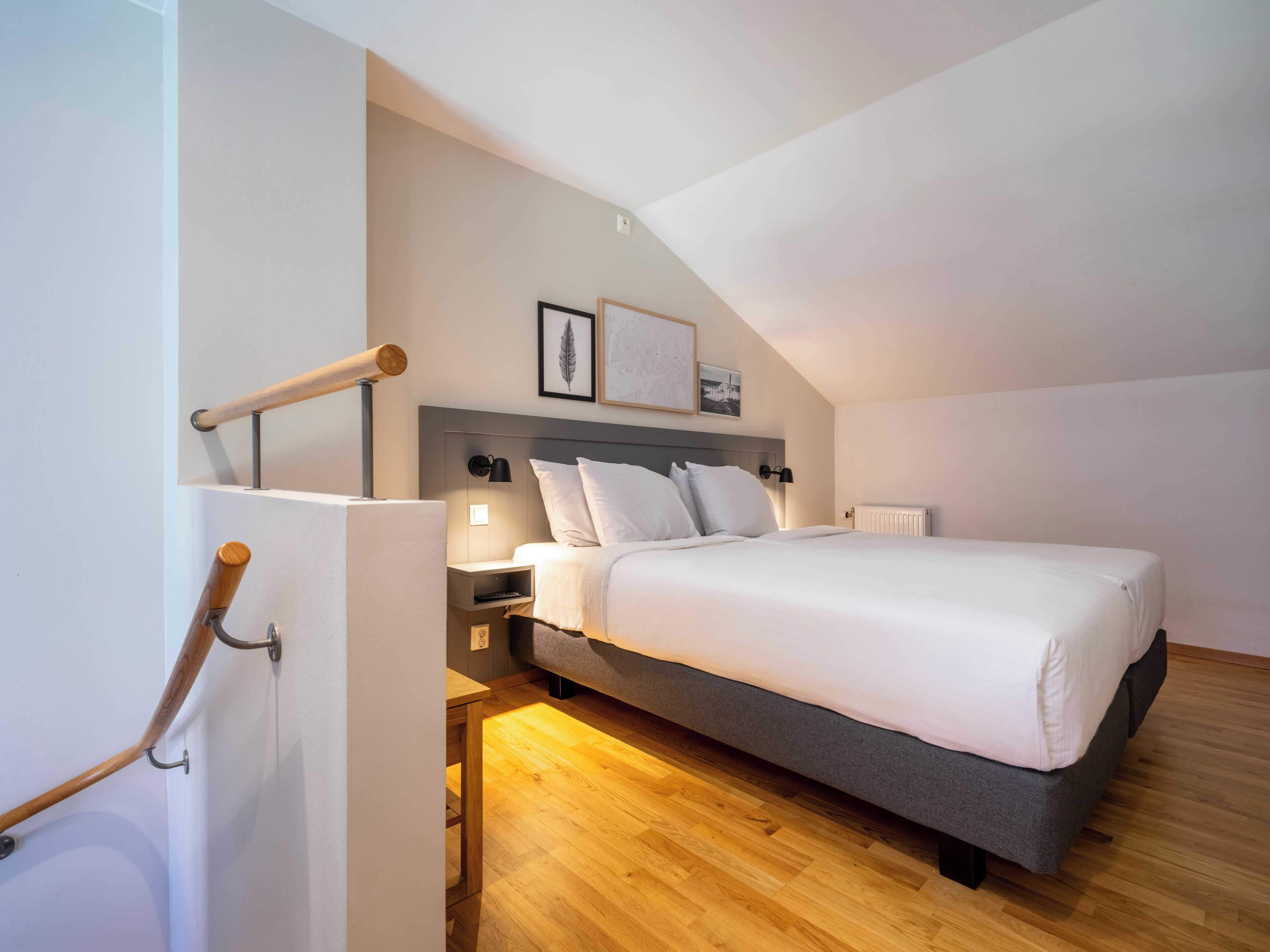 Quarto com piso de madeira, cama com roupa de cama branca, mesa de cabeceira, luminária na parede e quadro emoldurado.