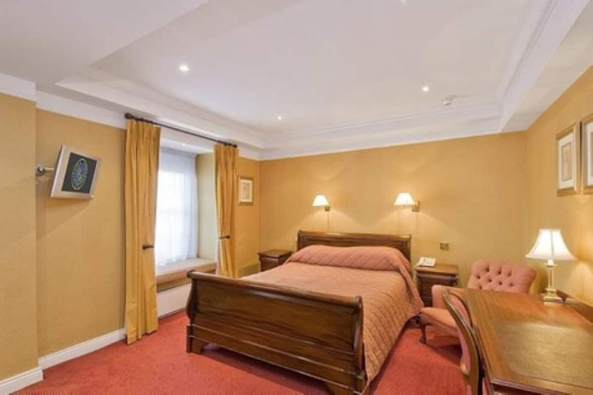 Quarto de hotel com cama de madeira, duas mesas de cabeceira, uma cadeira e uma janela com cortinas.