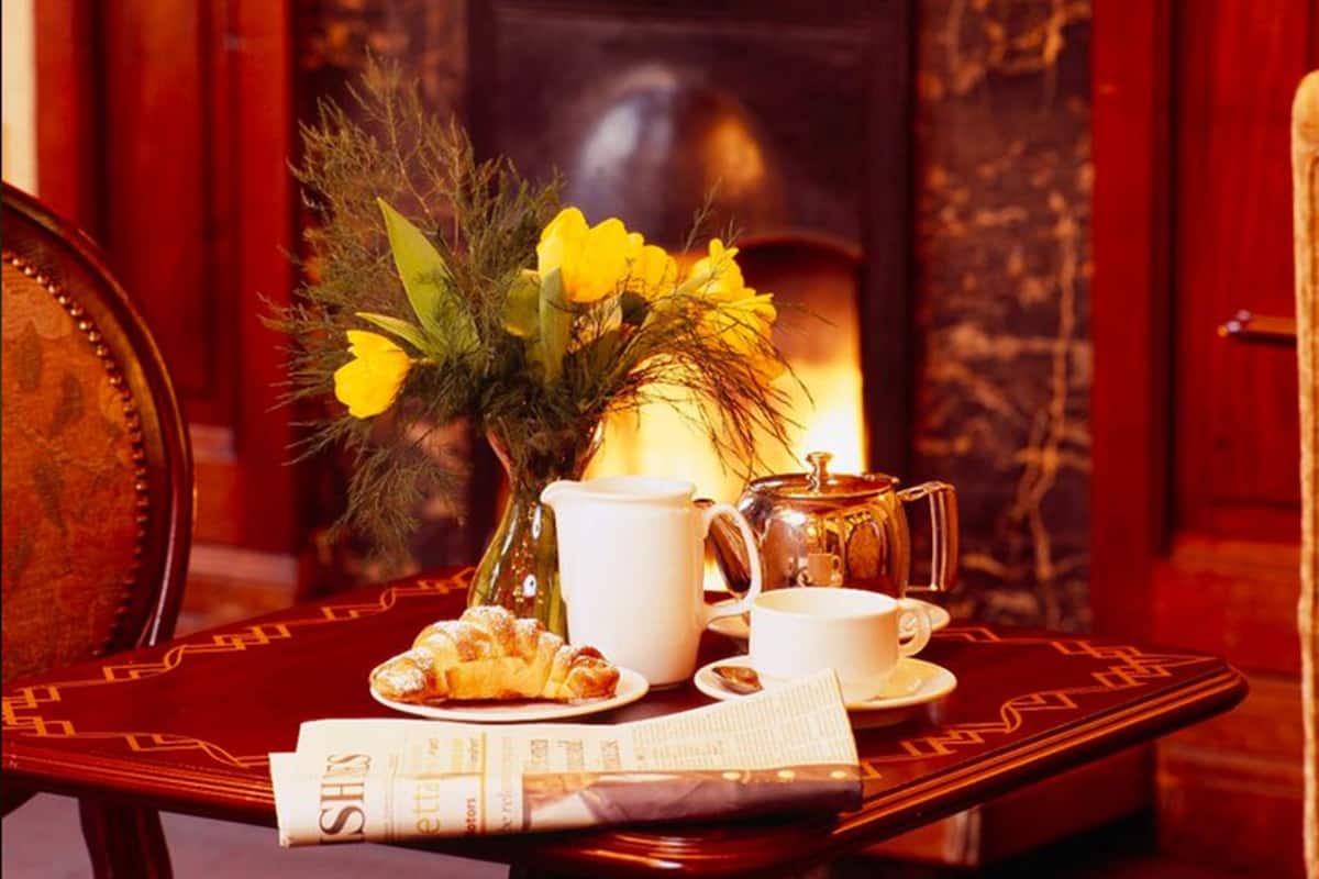 Uma mesa com jornal, um bule, xícaras, um croissant e um vaso com flores amarelas.