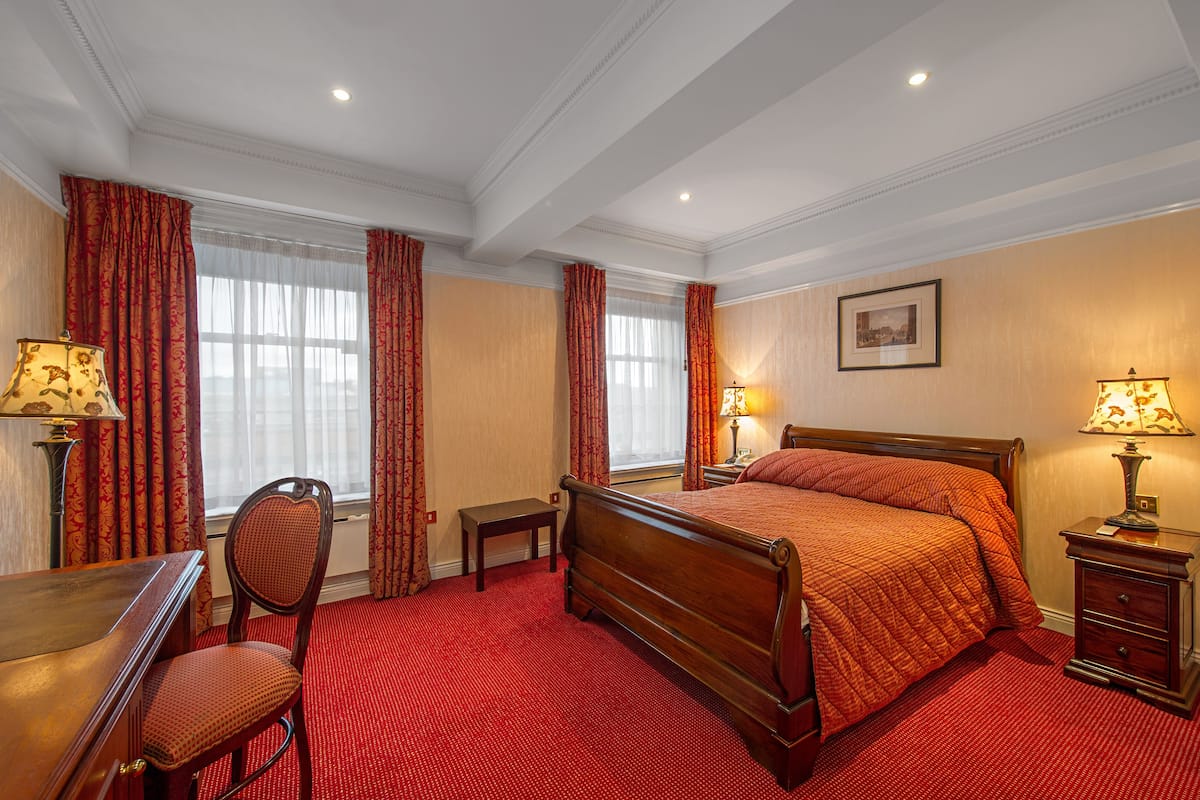 Quarto de hotel com cama de madeira, tapete vermelho, duas mesas de cabeceira com abajures, uma cadeira e uma janela com cortinas.