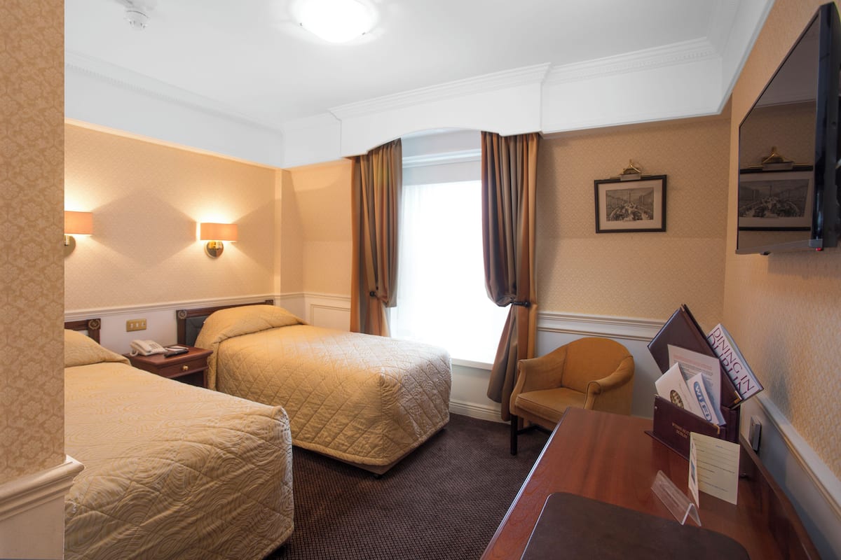 Quarto de hotel com duas camas, uma escrivaninha, uma cadeira, uma televisão e uma janela com cortinas.