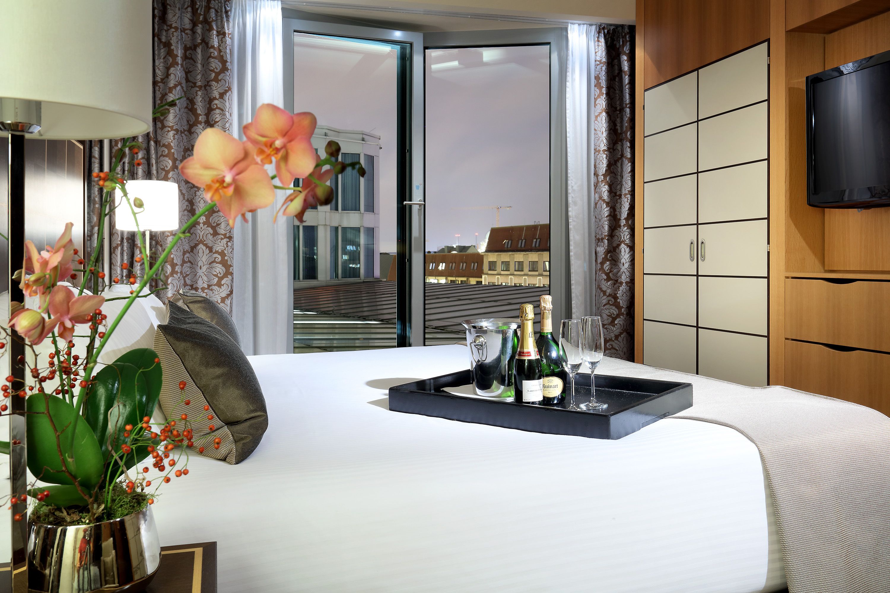Quarto de hotel com cama, bandeja com garrafas de champanhe e copos, vaso com flores e televisão na parede.