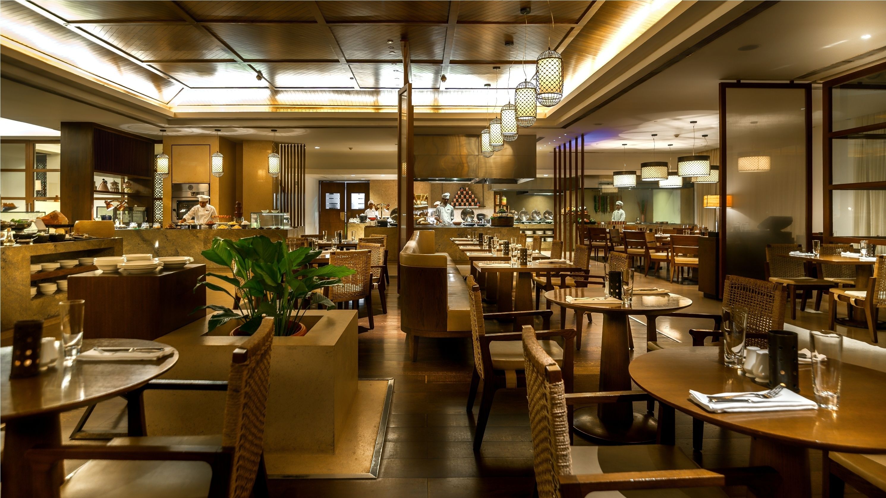 Interior de restaurante com mesas e cadeiras de madeira, área central de serviço e luminárias pendentes.