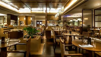 Interior de restaurante com mesas e cadeiras de madeira, área central de serviço e luminárias pendentes.