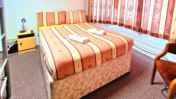 Quarto de hotel com cama, mesa de cabeceira, cadeira, escrivaninha e janela com cortinas.
