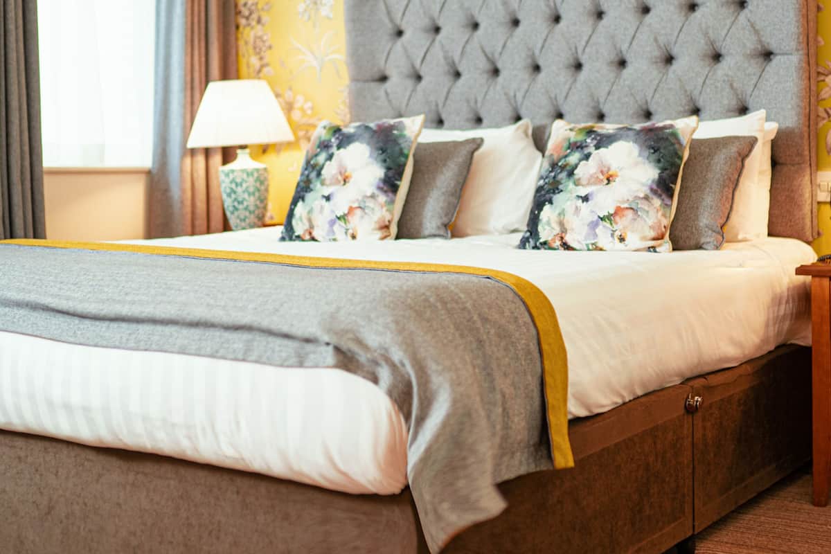 Cama bem arrumada com cabeceira cinza, almofadas florais e colcha listrada em amarelo.