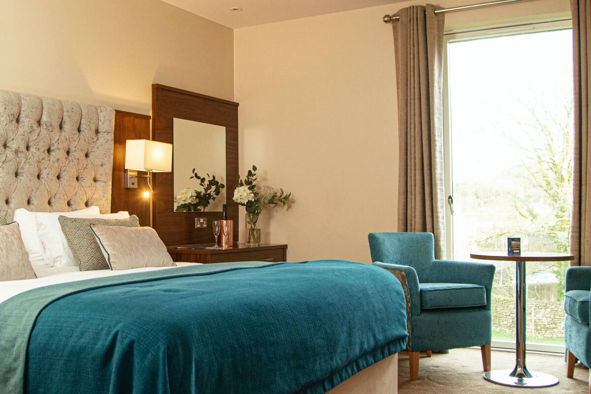 Quarto de hotel com uma cama grande, um espaço de estar com duas cadeiras, uma mesa pequena, um espelho e uma janela com cortinas.