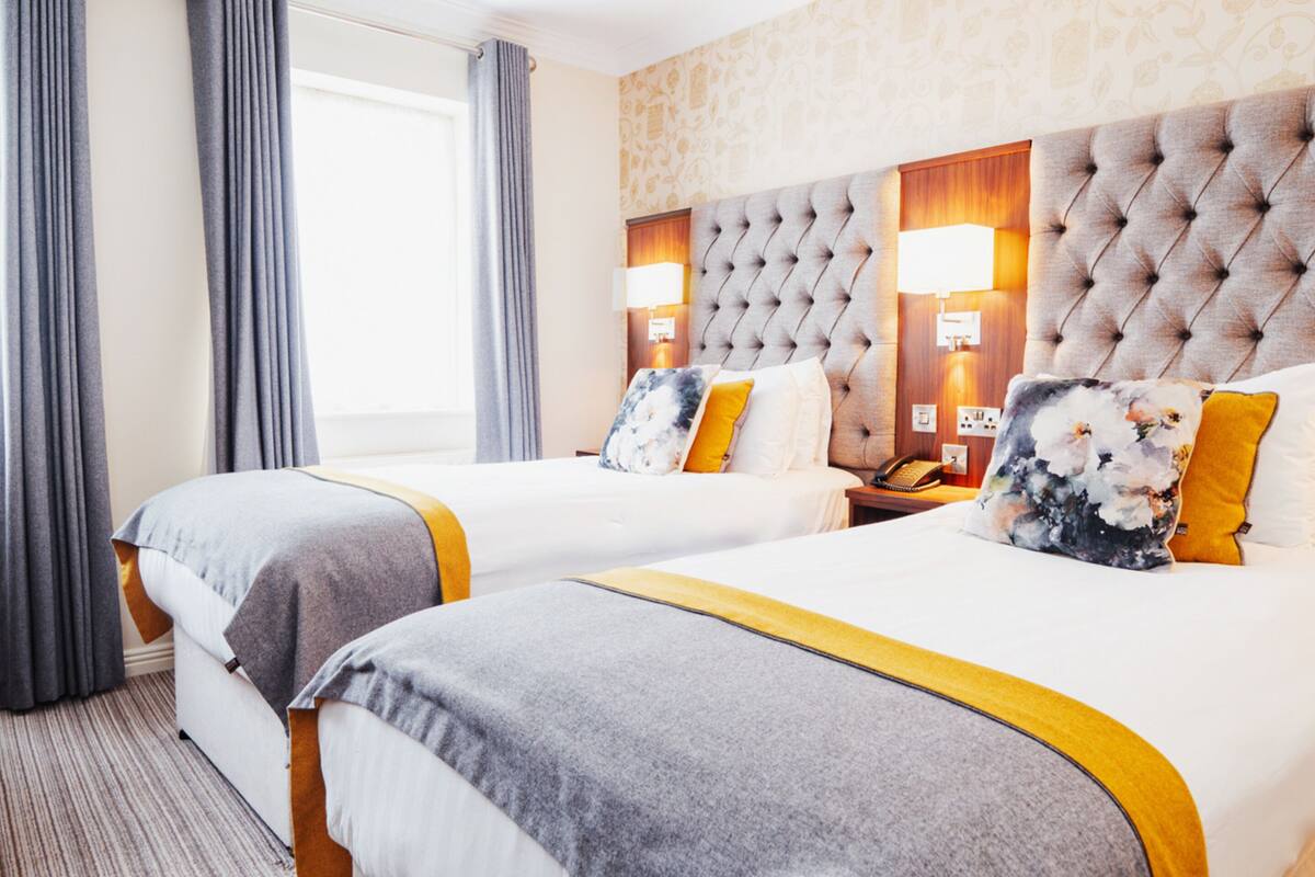 Quarto de hotel com duas camas, roupa de cama cinza e amarela, cabeceira estofada, luminárias laterais e papel de parede estampado.