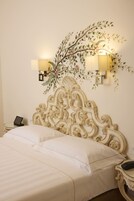 Quarto de hotel com uma cama grande e elaborada, uma mesa de cabeceira com abajur, e arandelas na parede com design de galho decorativo.