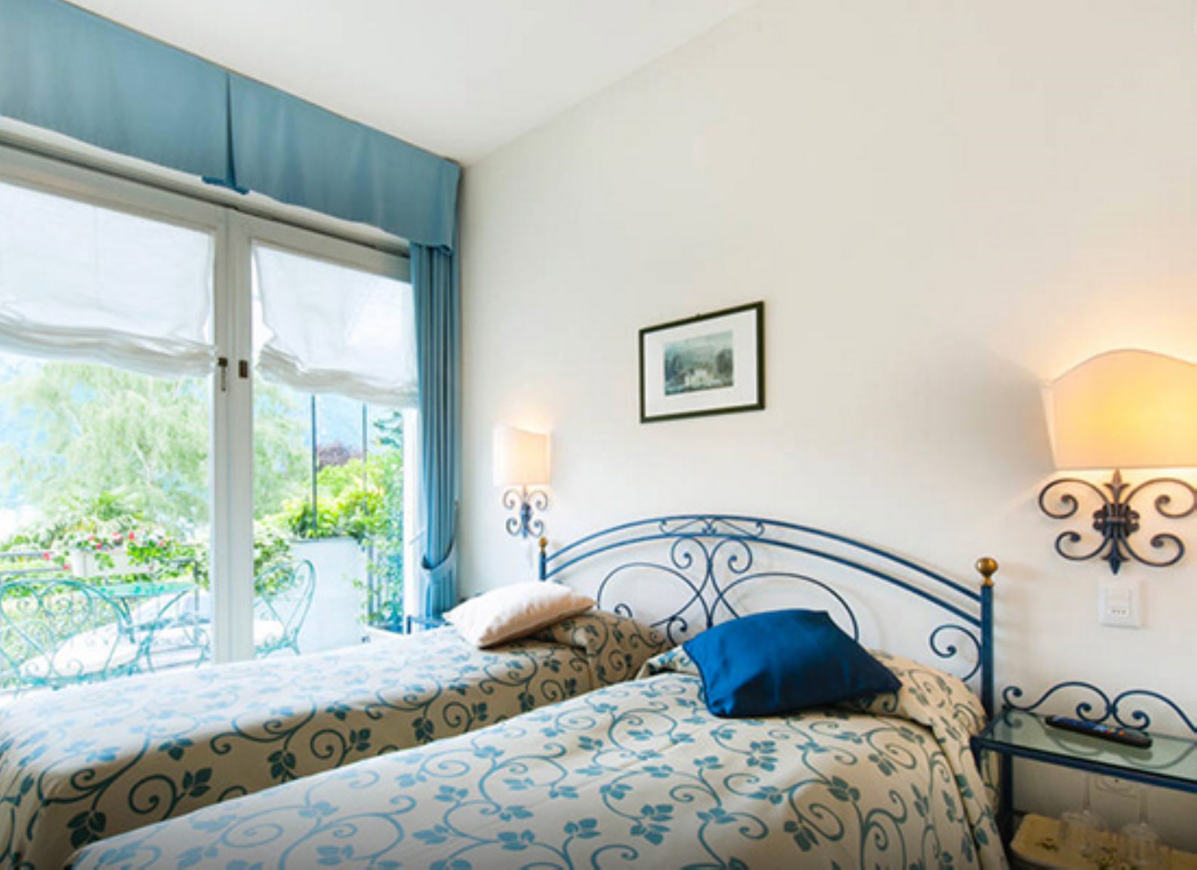 Quarto com duas camas, janela com cortinas, mesa de cabeceira com abajur e um quadro na parede.