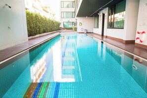 Piscina com piso de azulejos, adjacente a um edifício com varanda e um sebedo.