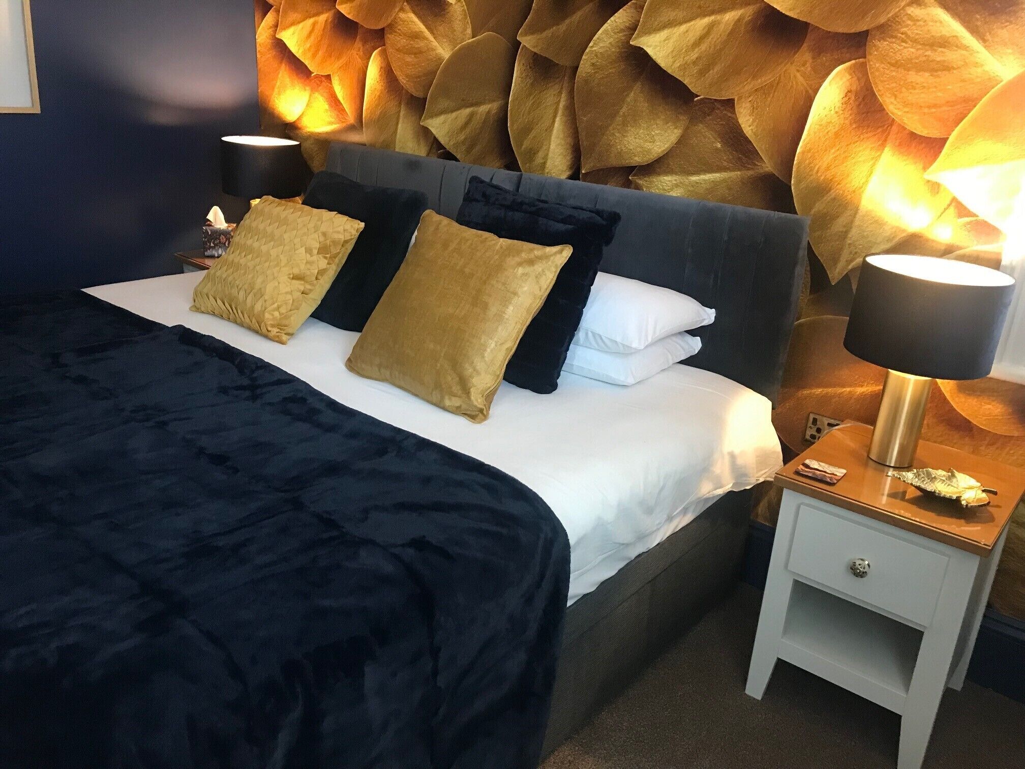 Cama bem arrumada com colcha azul escura e almofadas douradas e pretas, mesa de cabeceira com abajur e parede com design texturizado em formato de folhas.