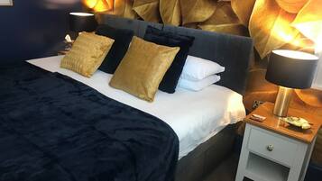 Cama bem arrumada com colcha azul escura e almofadas douradas e pretas, mesa de cabeceira com abajur e parede com design texturizado em formato de folhas.