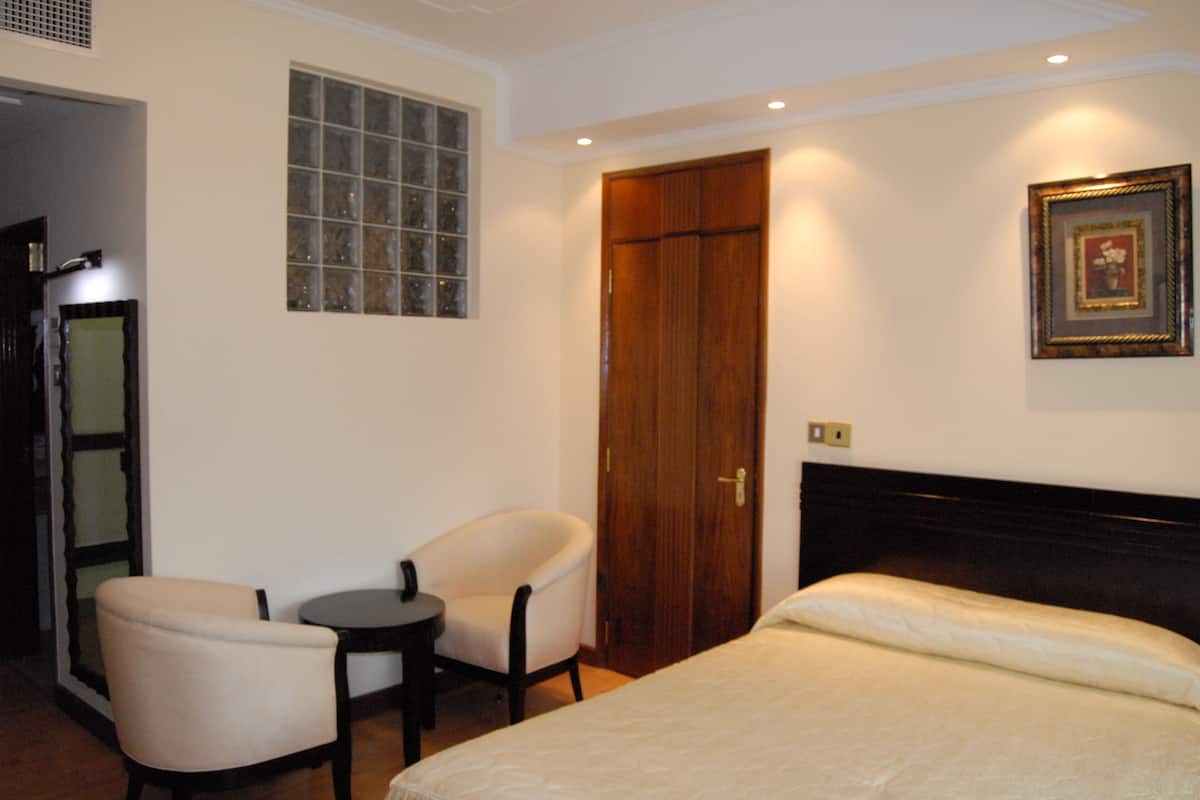 Quarto de hotel com uma cama, duas cadeiras, uma mesa pequena, uma porta de madeira e um quadro na parede.