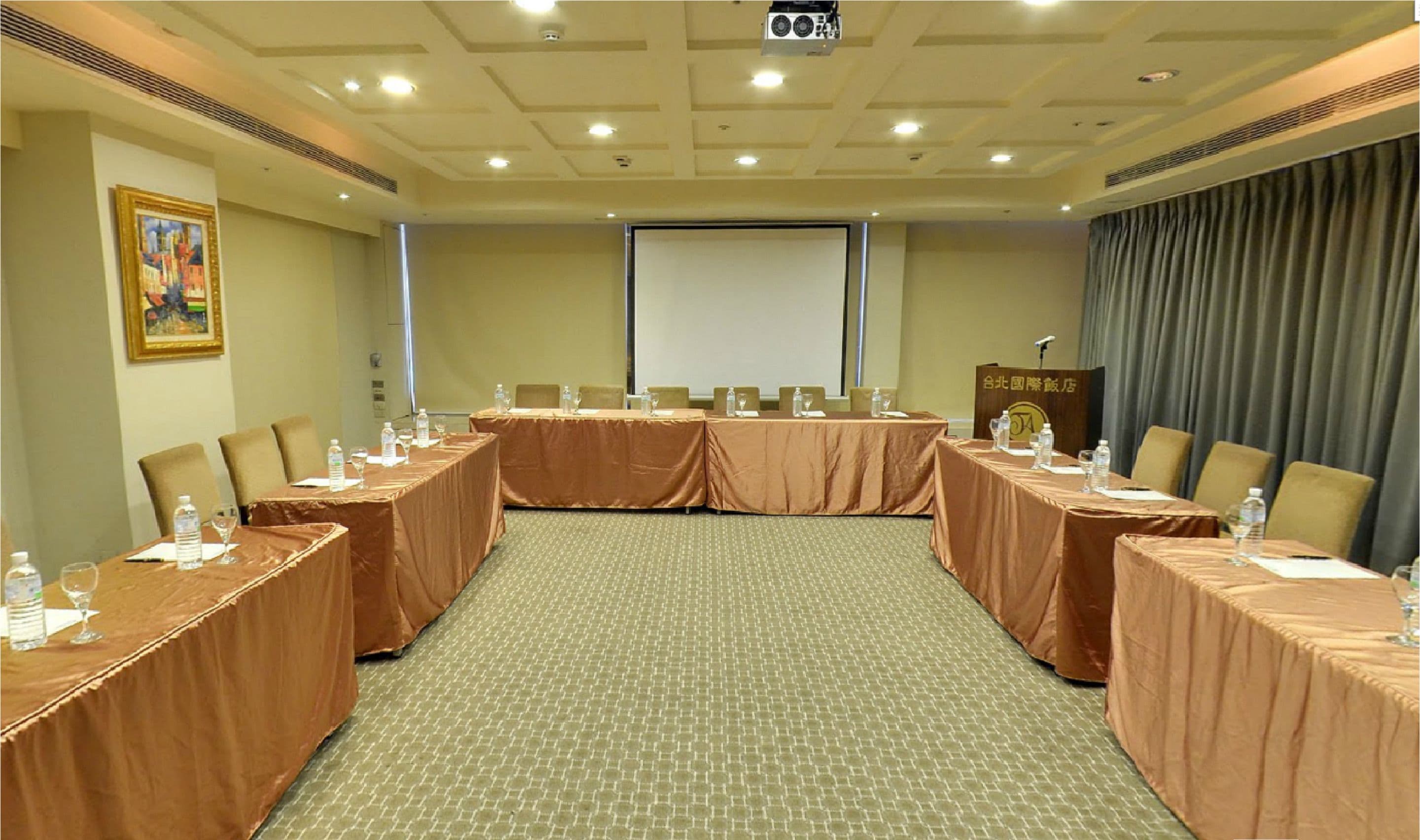 Sala de conferências com uma mesa comprida, cadeiras, tela de projetor e um púlpito.