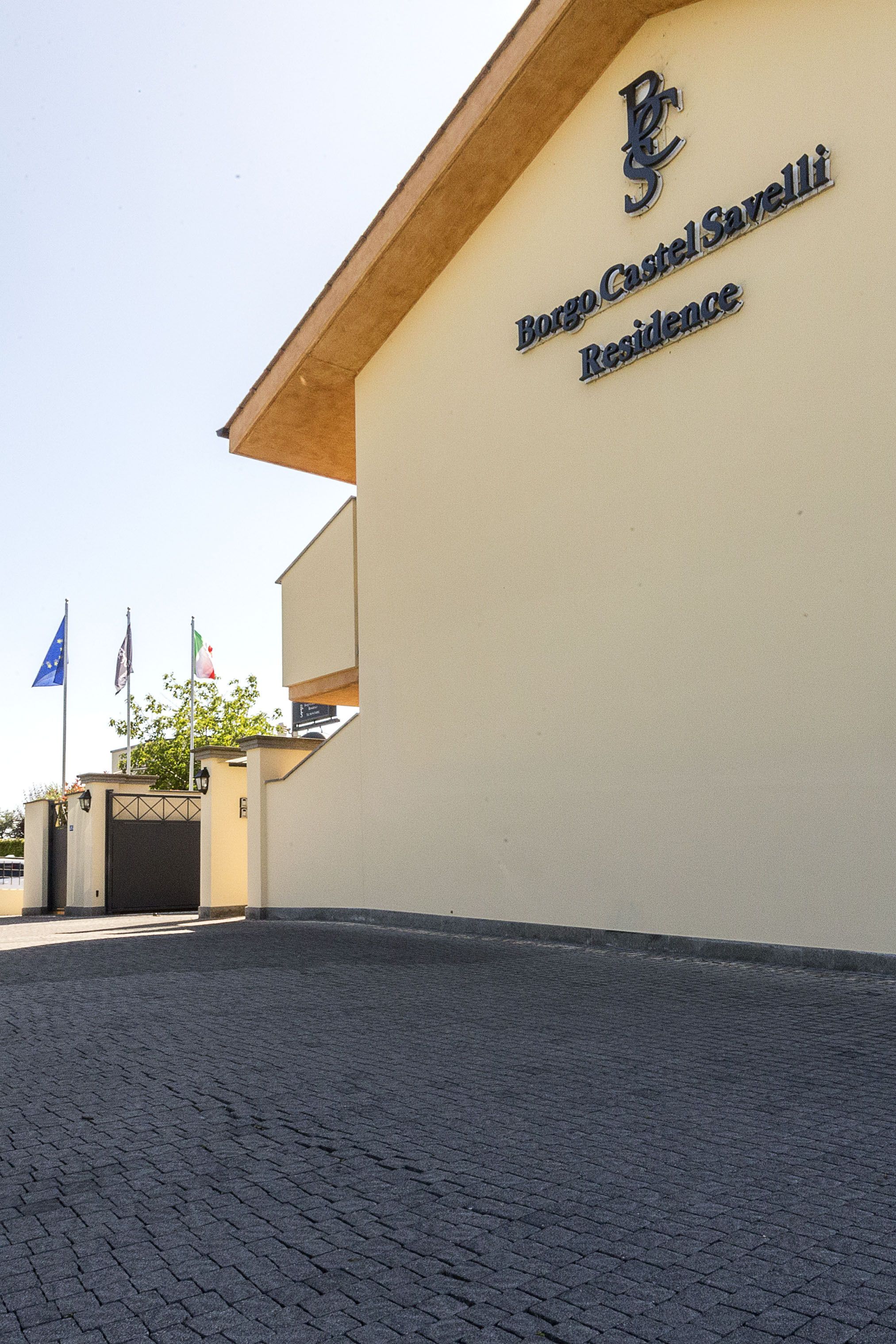 Exterior do Borgo Castel Savelli Residence, com entrada pavimentada e bandeiras.